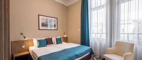 Quarto de hotel com uma cama grande, uma cadeira, uma mesinha de cabeceira, um abajur e um quadro na parede.
