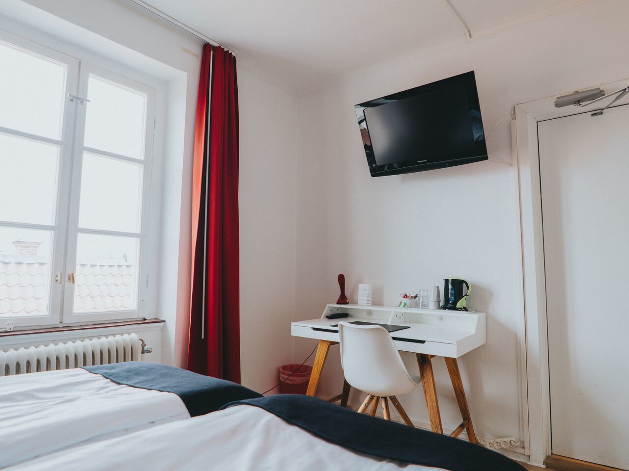 Quarto de hotel com cama, escrivaninha e cadeira, televisão na parede e janela com cortinas.