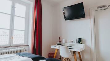 Quarto de hotel com cama, escrivaninha e cadeira, televisão na parede e janela com cortinas.