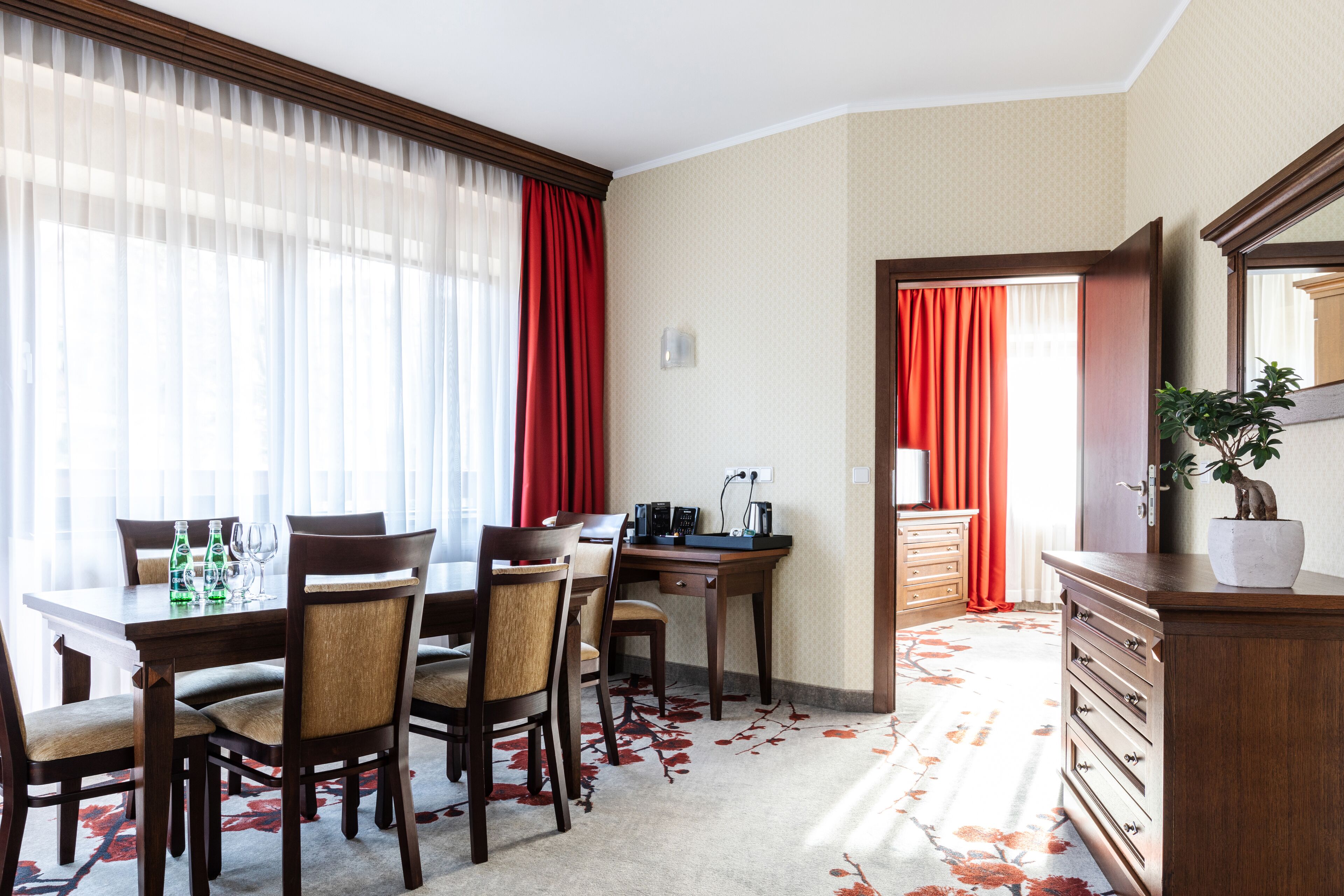 Quarto de hotel com área de refeições, uma escrivaninha com computador, cômoda e planta.