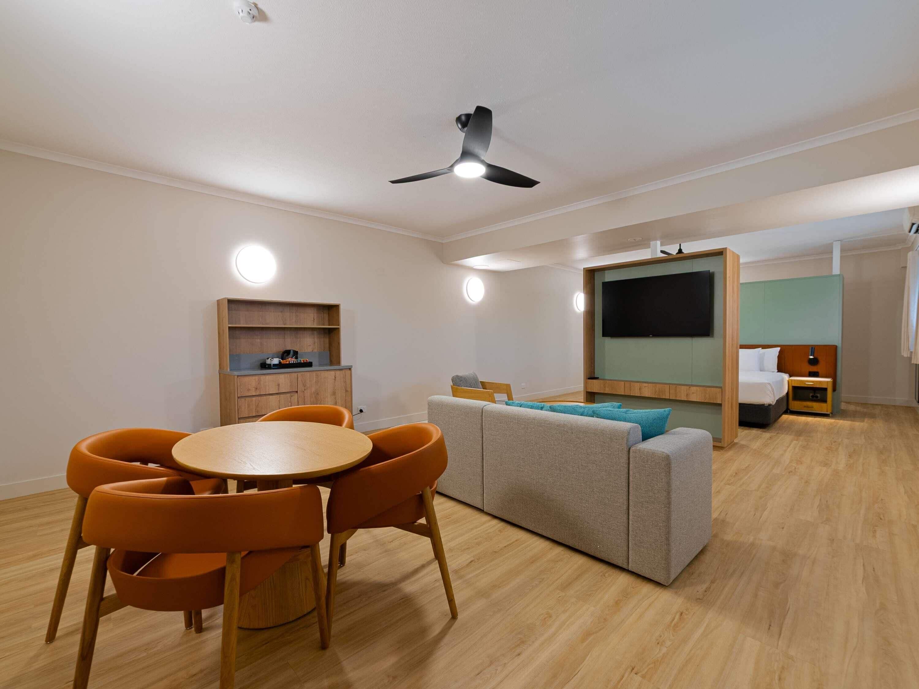 Quarto de hotel moderno com área de refeições, sofá, televisão e armário de madeira.