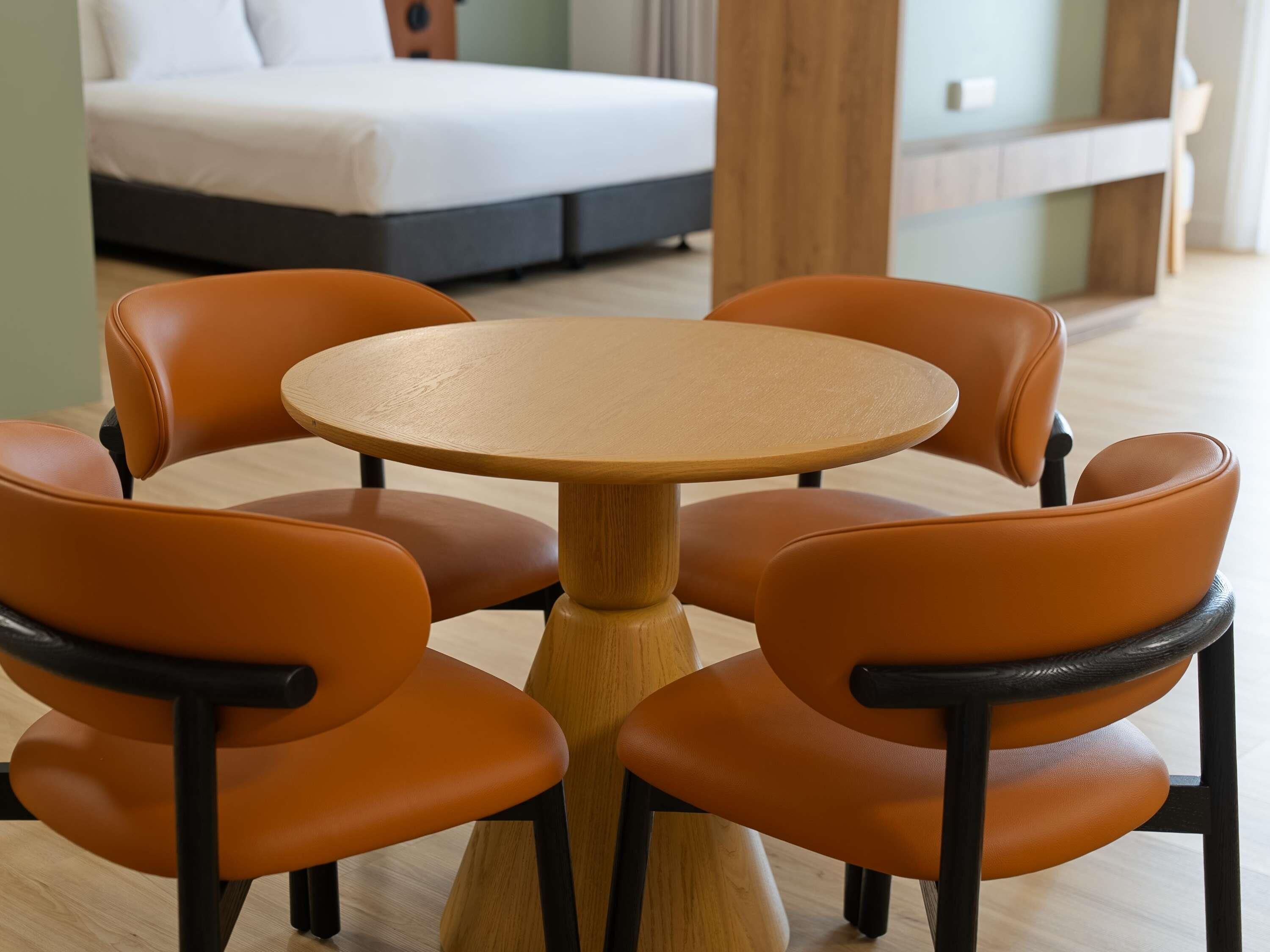 Um quarto com uma mesa redonda de madeira cercada por quatro cadeiras laranjas, uma cama ao fundo e um pilar de madeira.