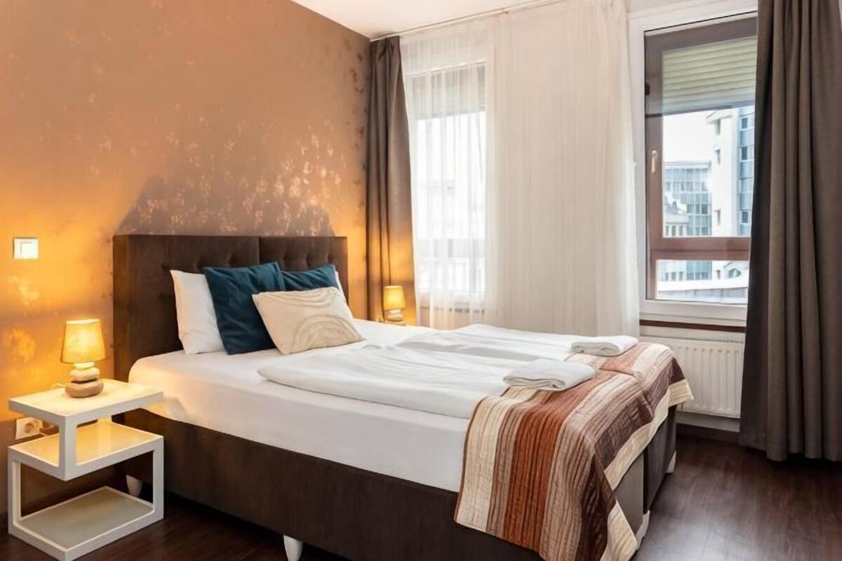 Quarto de hotel com uma cama grande, abajures, janela com cortinas e vista para prédios.