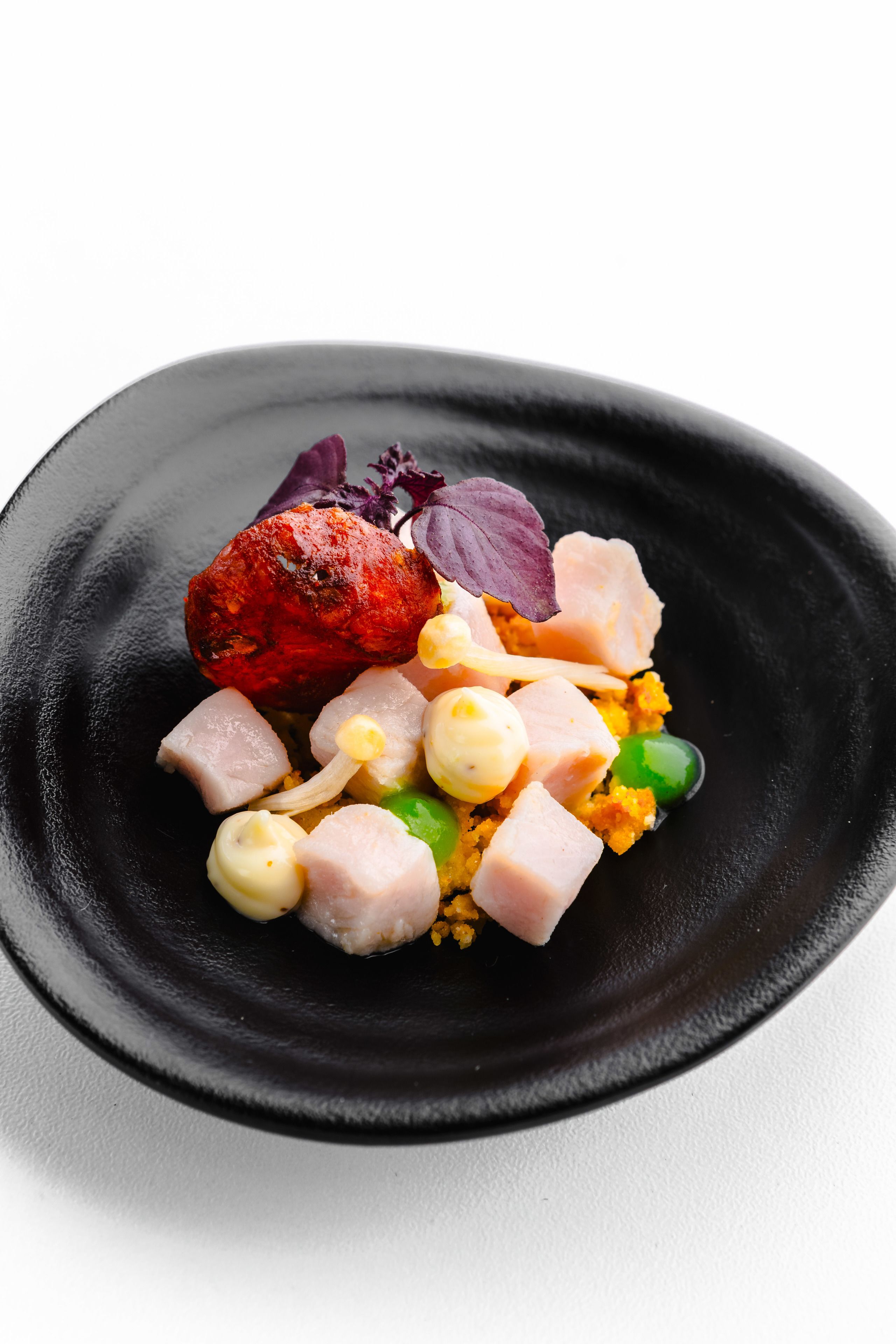 Um prato preto com diversos alimentos, incluindo um item vermelho, cubos de carne e vegetais verdes.