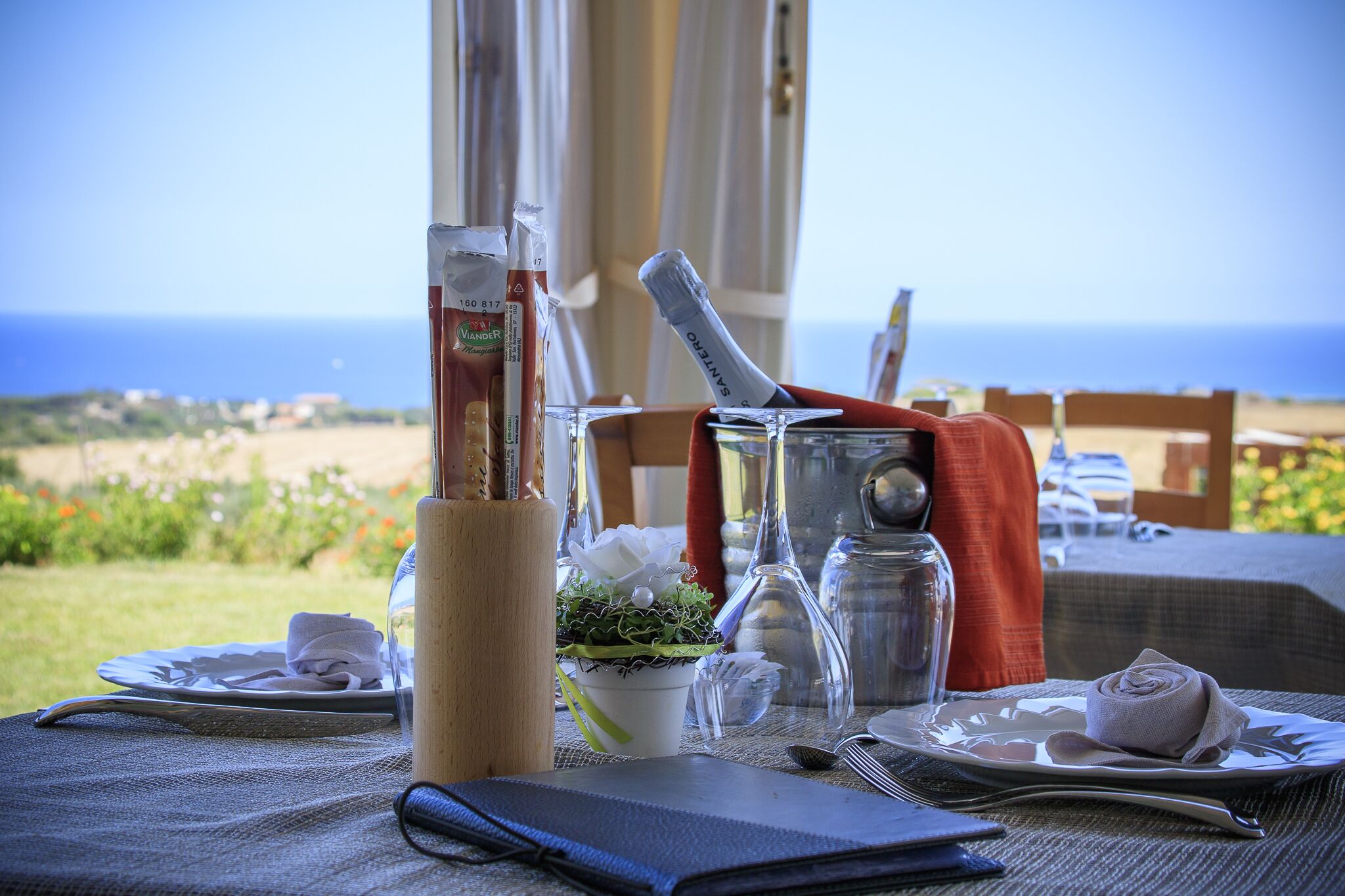 Mesa posta para dois com vista para o mar, uma garrafa de vinho, um jarro de vidro e um pequeno vaso com planta.