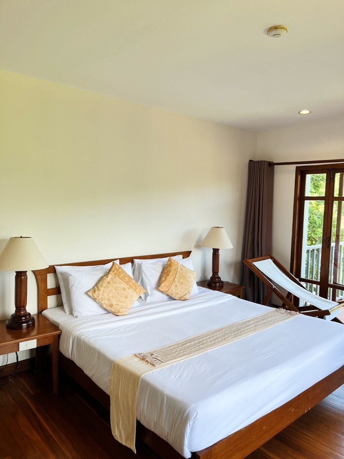 Uma cama bem arrumada com lençóis brancos e almofadas decorativas, cabeceira de madeira, duas luminárias de cabeceira e uma janela com vista para o verde.