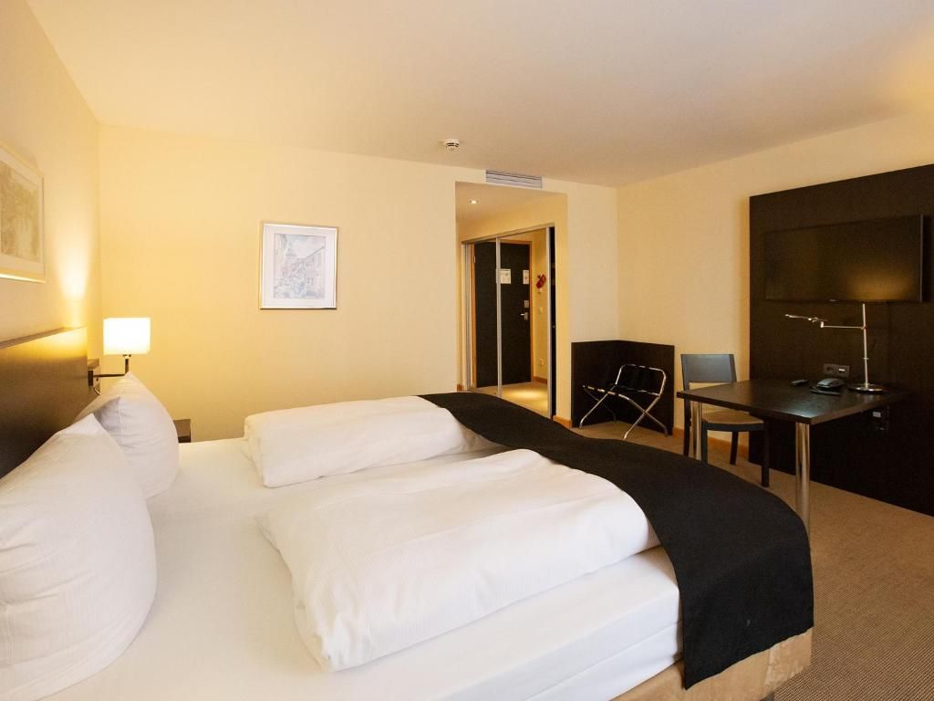 Quarto de hotel com cama, escrivaninha, cadeira e televisão de tela plana.