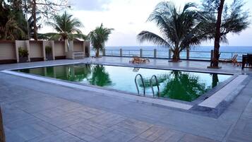 Piscina com vista para o mar, cercada por palmeiras e cadeiras de descanso.