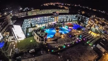 Um hotel grande e iluminado, com área de piscina central, rodeado por um jardim paisagístico e um estacionamento.