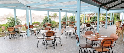 Um café ao ar livre com mesas e cadeiras dispostas sob um toldo azul.