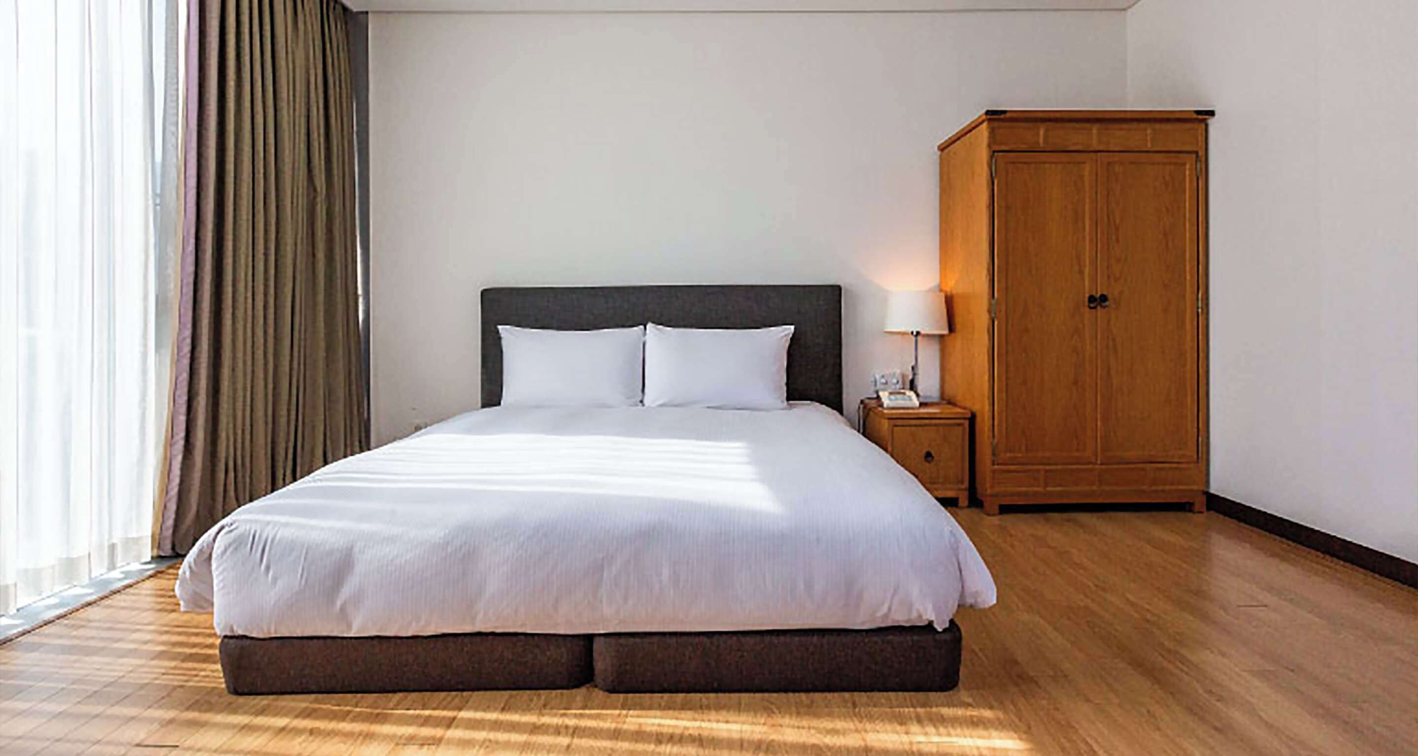 Quarto de hotel com uma cama grande, um armário de madeira, uma mesa de cabeceira com abajur e uma janela com cortinas.