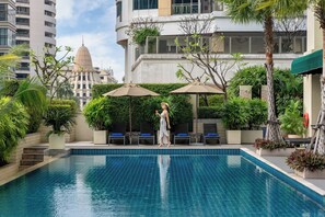 Piscina com água azul cristalina, cercada por espreguiçadeiras e guarda-sóis, com paisagem urbana ao fundo.
