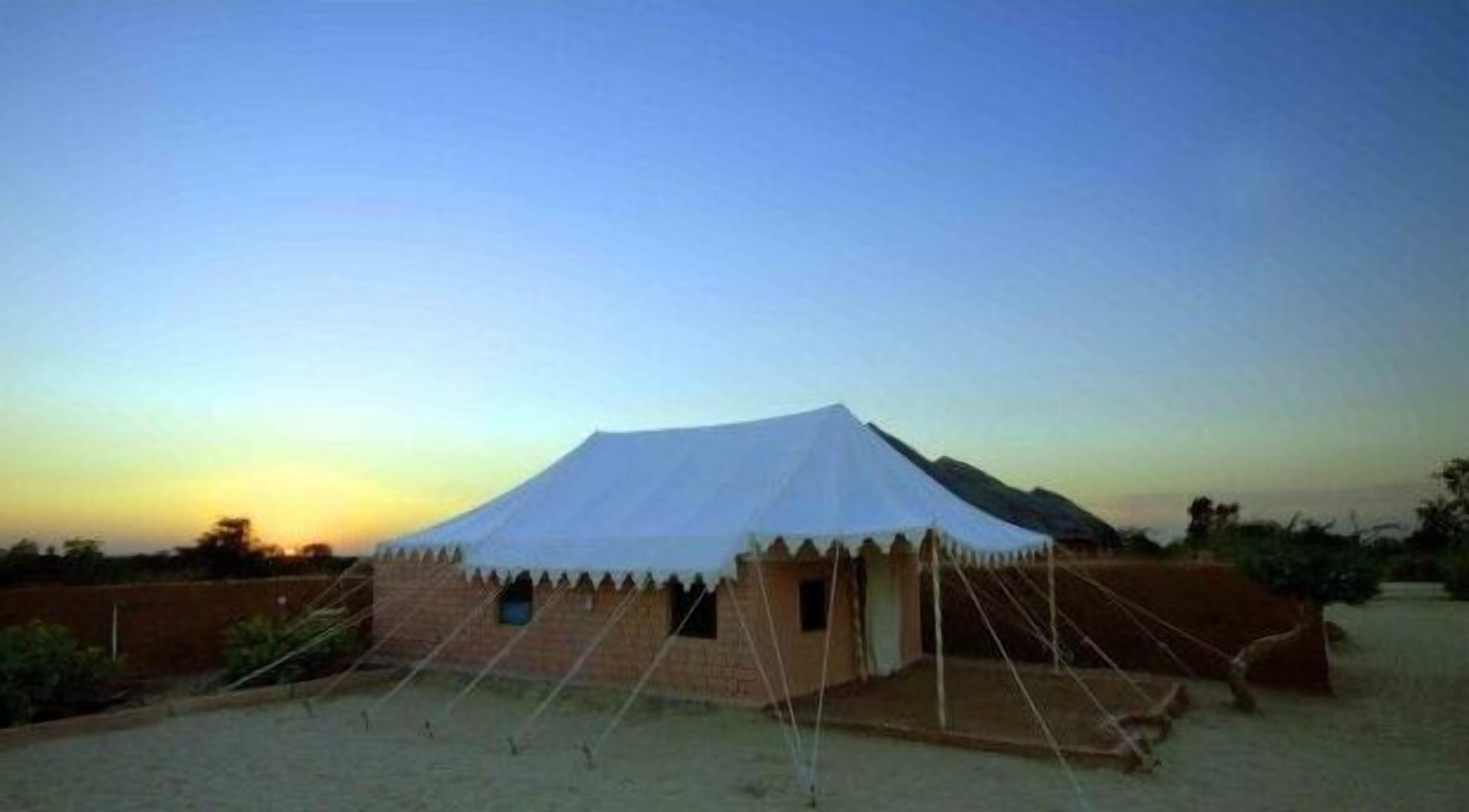 Uma tenda grande com teto em forma de pirâmide e pequena entrada, rodeada por uma estrutura de tijolos e uma área arenosa.