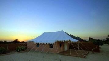 Uma tenda grande com teto em forma de pirâmide e pequena entrada, rodeada por uma estrutura de tijolos e uma área arenosa.