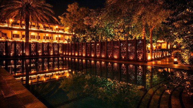 Vista noturna de um hotel com piscina refletindo luzes, uma palmeira e uma cerca decorativa.