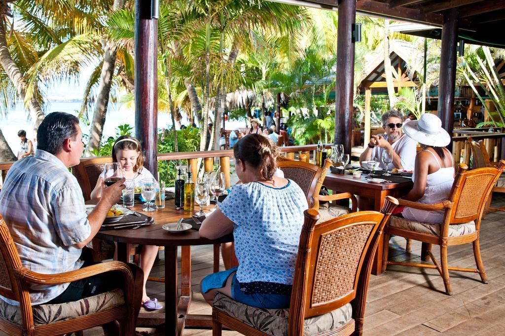 Um grupo de pessoas jantando em um restaurante ao ar livre, com móveis de madeira e um cenário tropical.