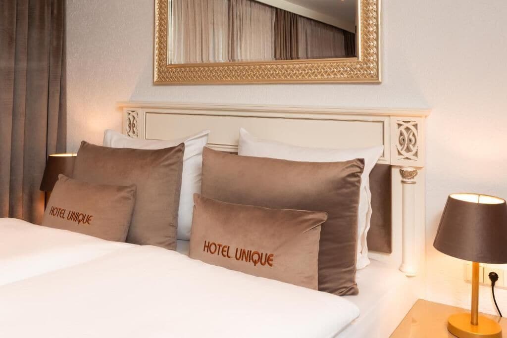 Quarto de hotel com cama, almofadas, mesa de cabeceira, abajur e espelho na parede.