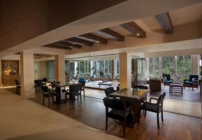 Um espaço amplo e interno com uma mesa grande para jogar xadrez, várias cadeiras e vista para o ginásio através de grandes janelas.