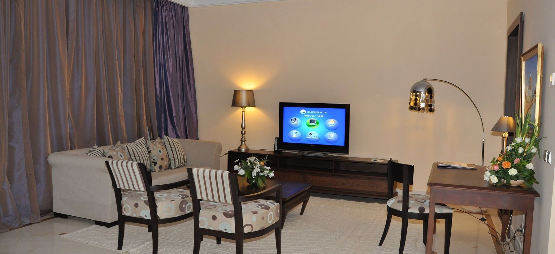 Sala de estar com sofá, duas poltronas, mesa de centro, televisão e escrivaninha com luminária e flores.
