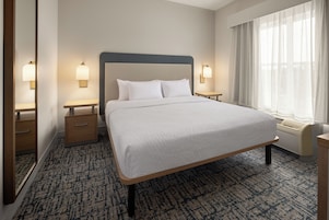Quarto de hotel com uma cama grande, duas mesas de cabeceira, um criado-mudo, uma janela com cortinas leves e uma luminária fixada na parede.