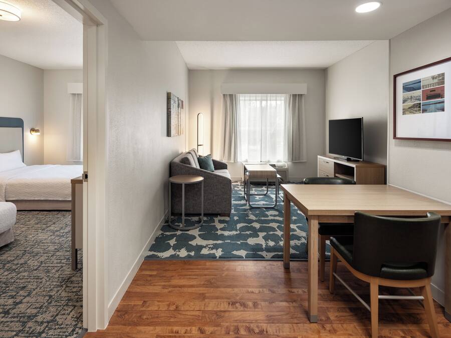Quarto de hotel com cama, sofá, mesa de jantar com cadeiras, televisão e uma janela grande com cortinas.