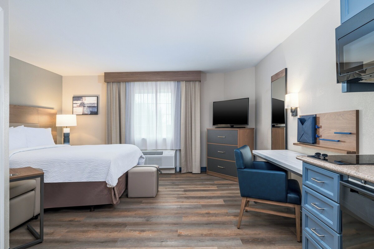 Quarto de hotel com cama, escrivaninha, cadeira, televisão e uma pequena cozinha.