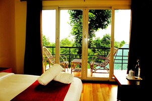 Quarto de hotel com cama, varanda com cadeiras de vime e vista para o verde lá fora.