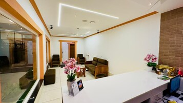 Uma área de recepção com balcão branco, espaço de estar com sofás, uma planta em vaso e um relógio fixado na parede.