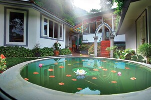 Um pátio tranquilo com um lago central, rodeado por vegetação exuberante e um edifício tradicional com telhado em azulejos.