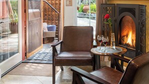 Um quarto acolhedor com lareira, uma poltrona confortável, uma mesinha com copos de vinho e vista para o exterior.