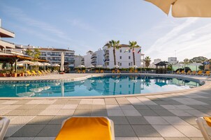 Uma área de piscina com espreguiçadeiras e guarda-sóis, cercada por edifícios com varandas e palmeiras.