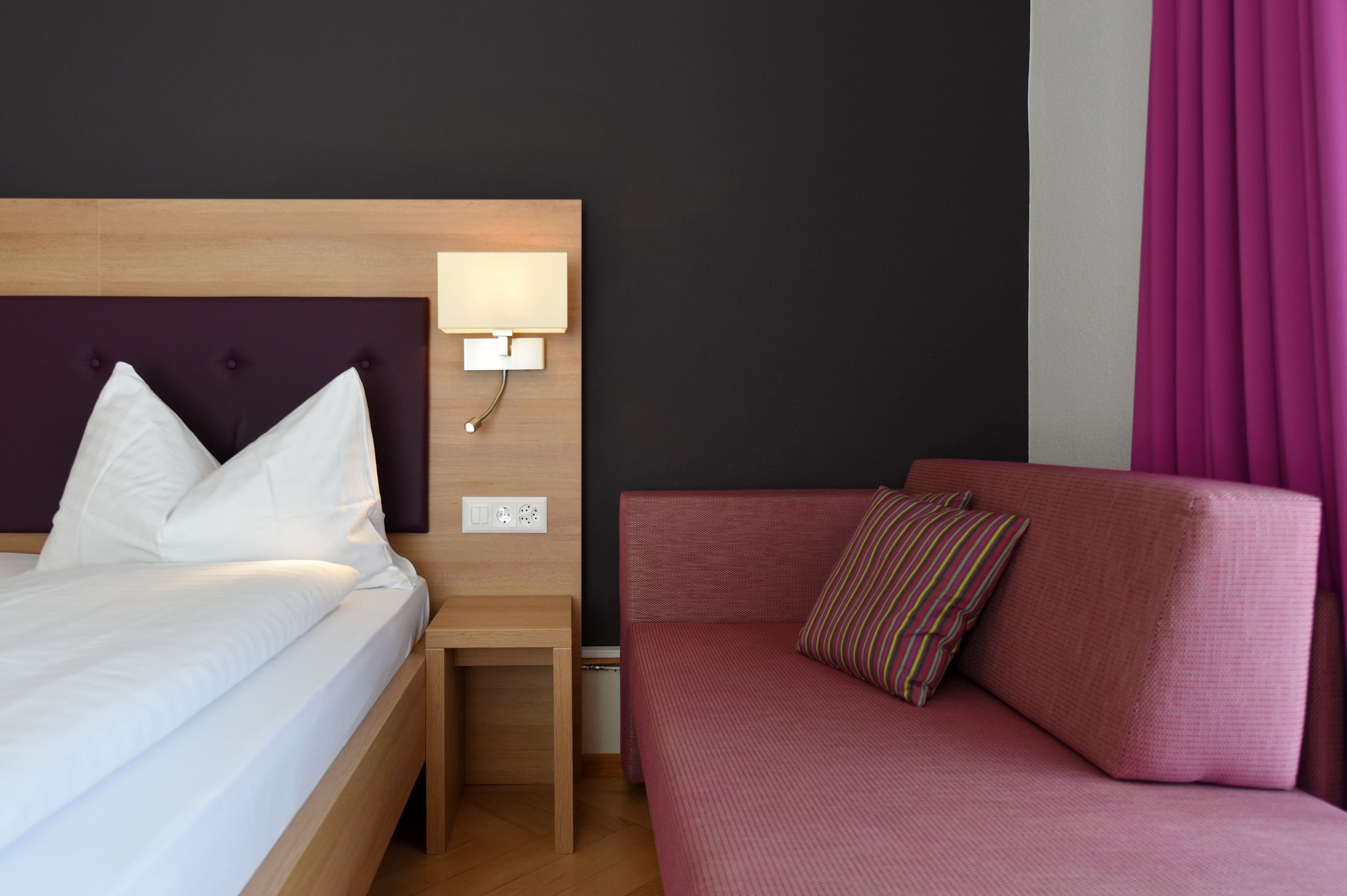 Quarto de hotel com cama, mesa de cabeceira, abajur e sofá.