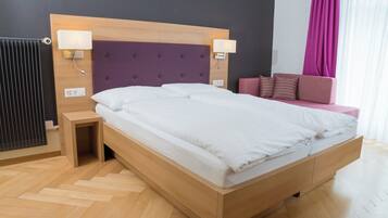 Quarto de hotel moderno com cama de madeira, duas mesas de cabeceira, painel de cabeceira e um pequeno espaço de estar com sofá rosa.