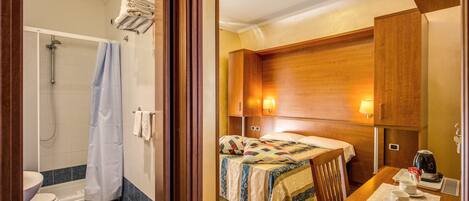 Quarto de hotel com chuveiro, vaso sanitário, cama, mesa de cabeceira e uma pequena mesa com um controle remoto.