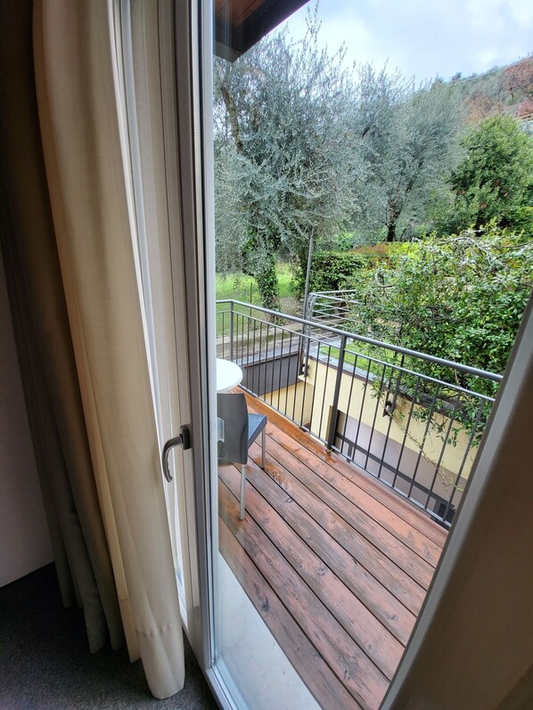 Uma varanda com deck de madeira, guarda-corpo de metal e vista para um jardim com árvores e arbustos.