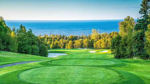 Um campo de golfe com lagoa, bunkers de areia e vista para o oceano.