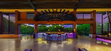 Uma área de bar ao ar livre com balcão central, assentos e vista para vegetação.