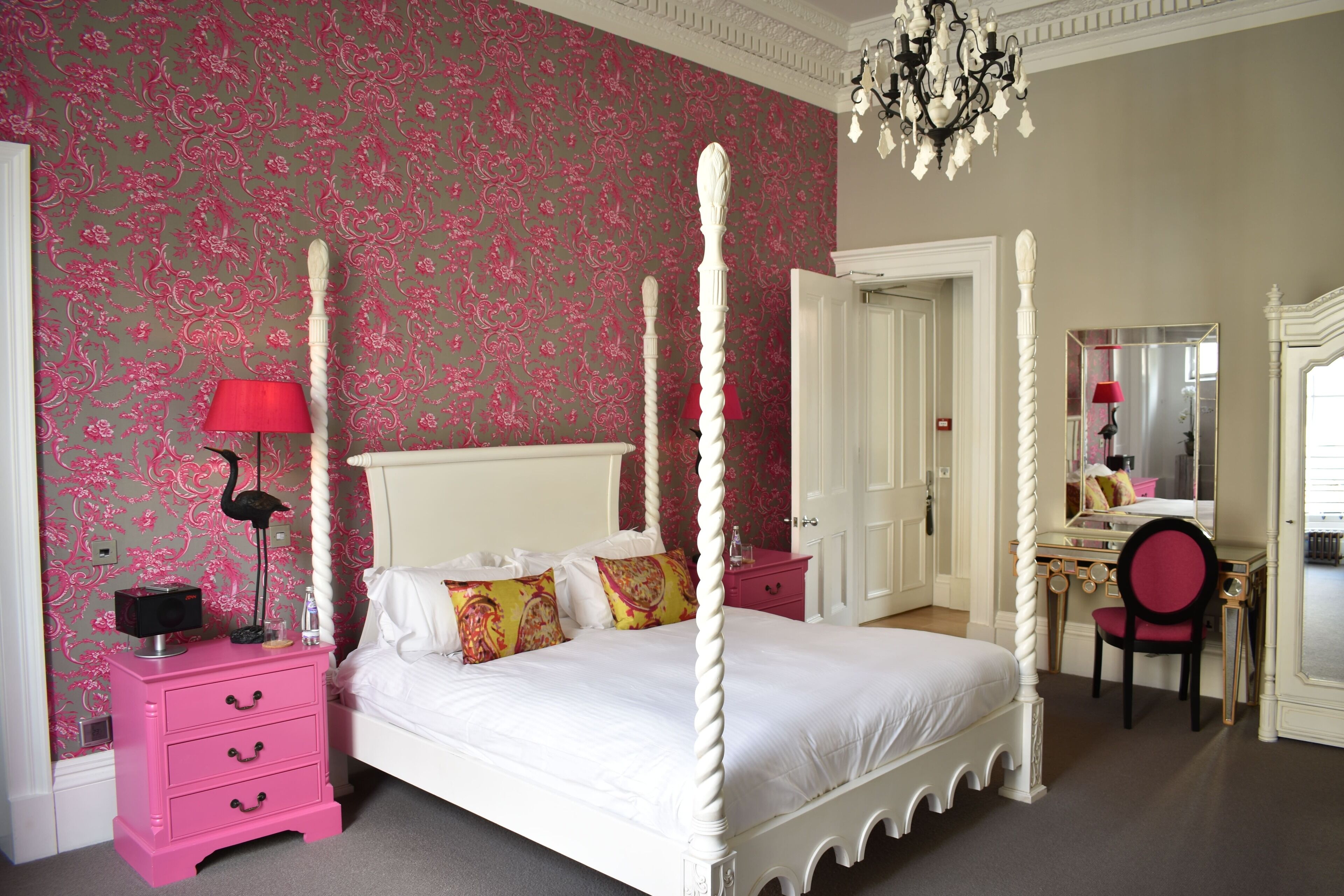 Uma cama de dossel com roupa de cama branca, uma mesa de cabeceira rosa, um abajur vermelho e um espelho em uma escrivaninha com uma cadeira vermelha.