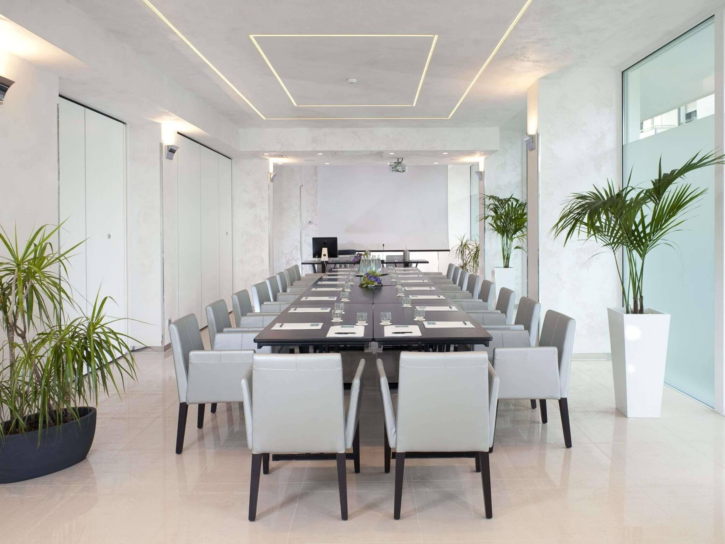 Uma sala de conferências moderna com uma mesa comprida, cadeiras, projetor e plantas em vasos.