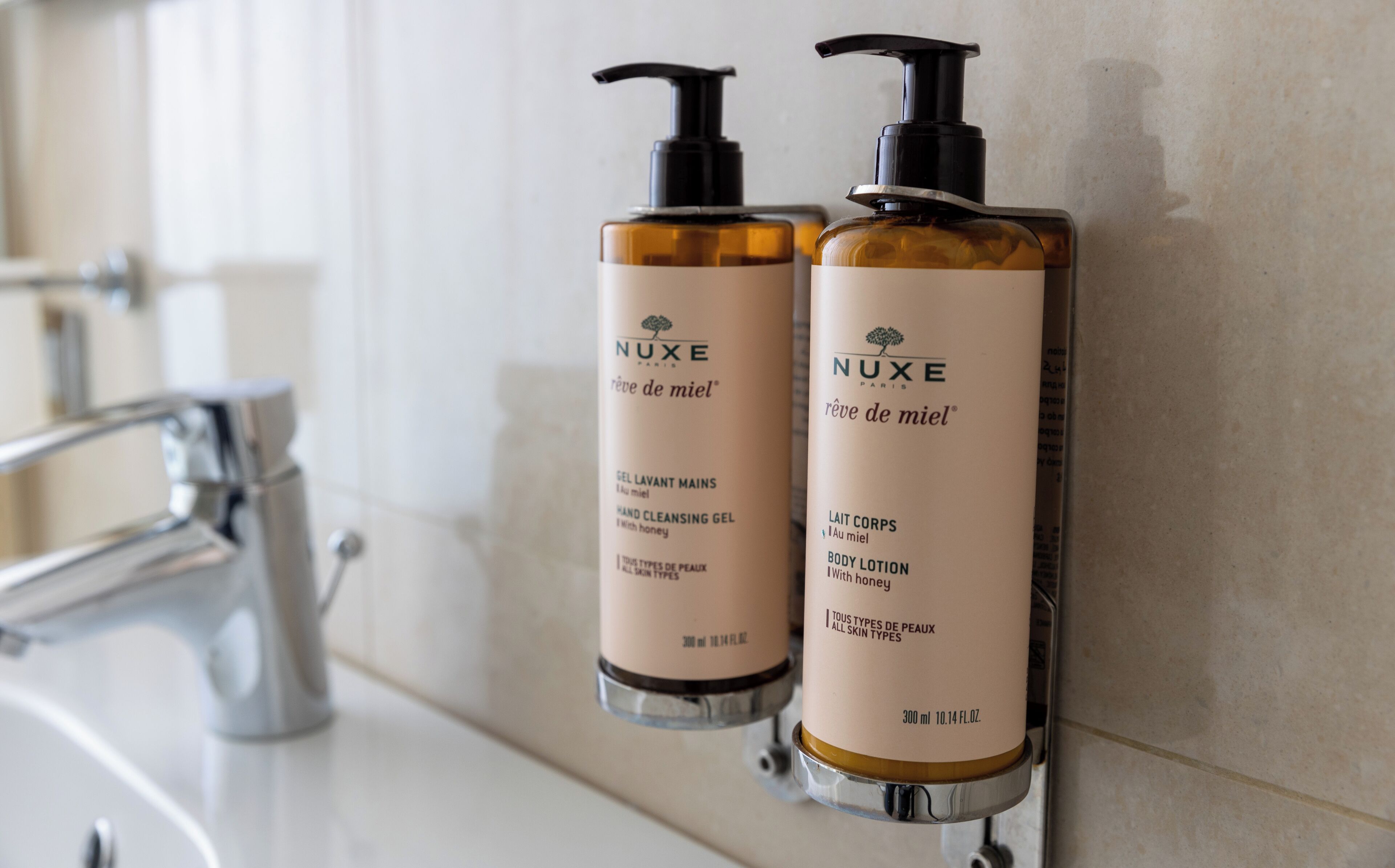 Duas embalagens de produtos de cuidados para a pele Nuxe Paris numa parede de banheiro.