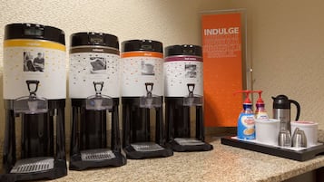 Quatro dispensadores de café identificados como tradicional, encorpado, descafeinado e água quente, juntamente com uma cafeteira, açúcar e leite, sobre uma bancada.