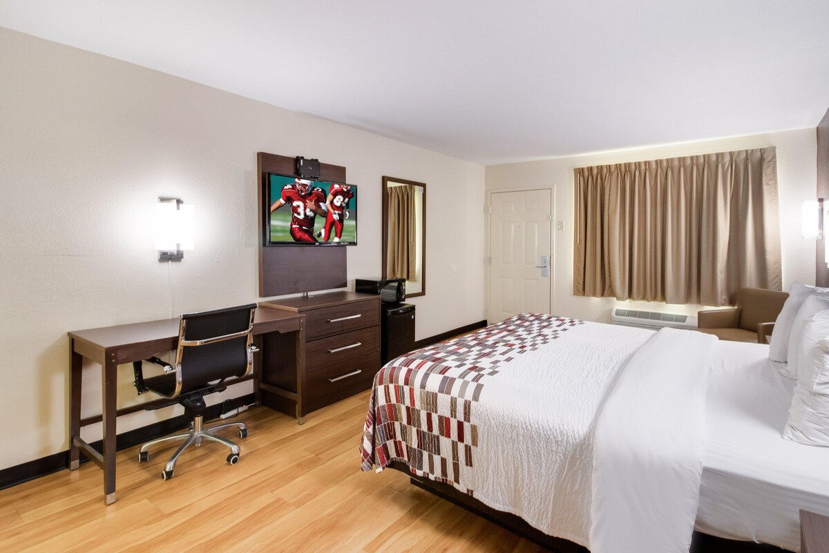 Quarto de hotel com cama, escrivaninha com cadeira, televisão e janela com cortinas.
