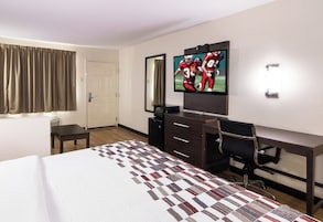 Quarto de hotel com cama, escrivaninha, cadeira, televisão e porta.