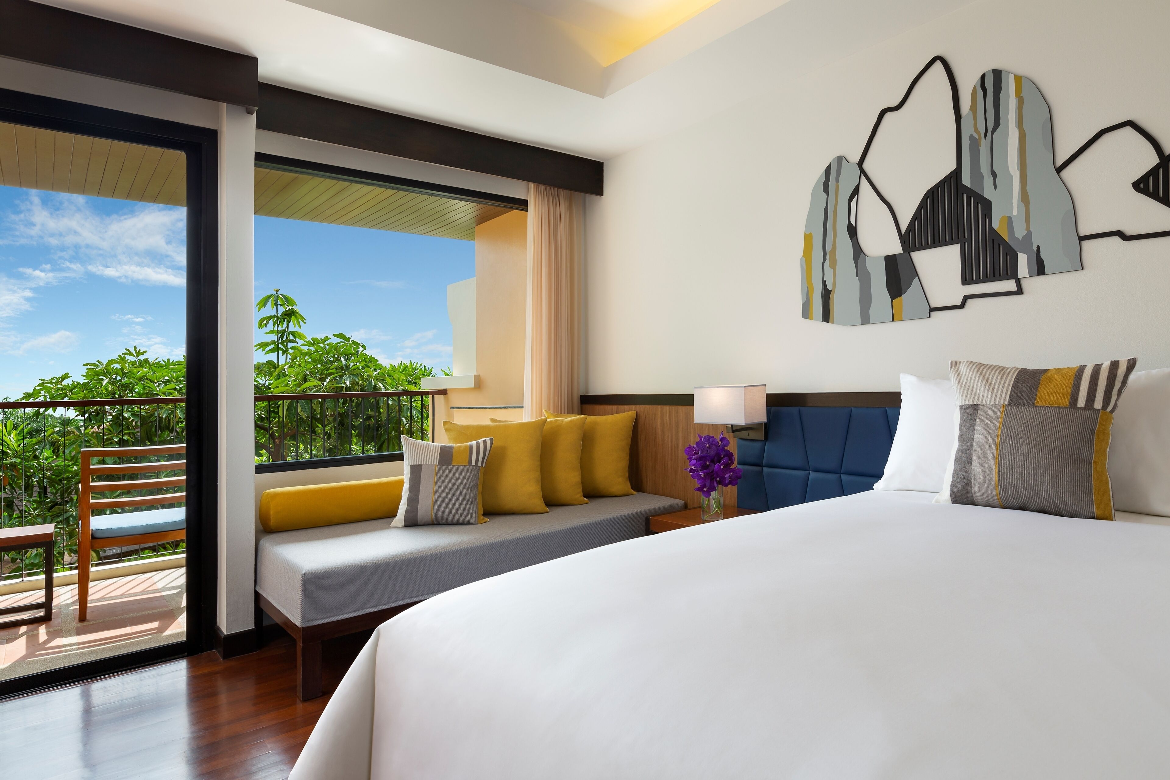 Quarto de hotel com uma cama grande, um sofá com almofadas amarelas, vista para área verde e uma obra de arte abstrata moderna na parede.