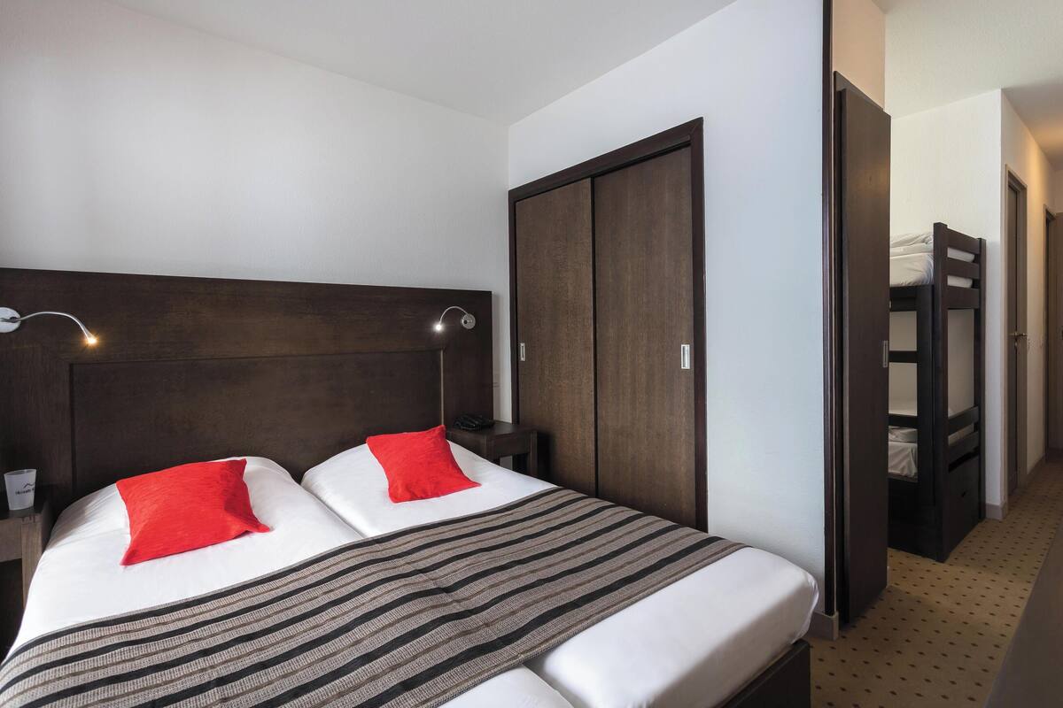 Quarto de hotel com uma cama grande, cabeceira de madeira, guarda-roupa e uma pequena mesa com abajur.