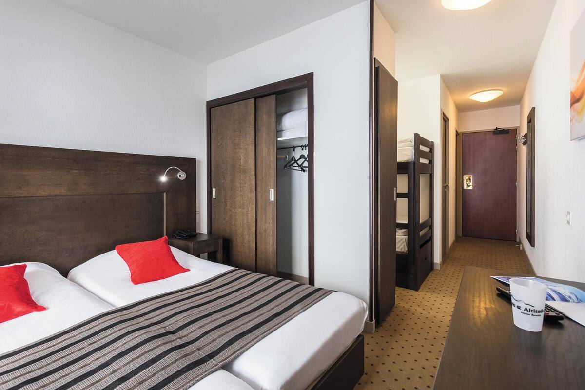 Quarto de hotel com cama, guarda-roupa, escrivaninha e porta que dá para outro cômodo.