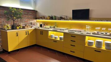Cozinha amarela e luminosa com parede de tijolos, uma televisão instalada acima do balcão e diversos alimentos expostos.