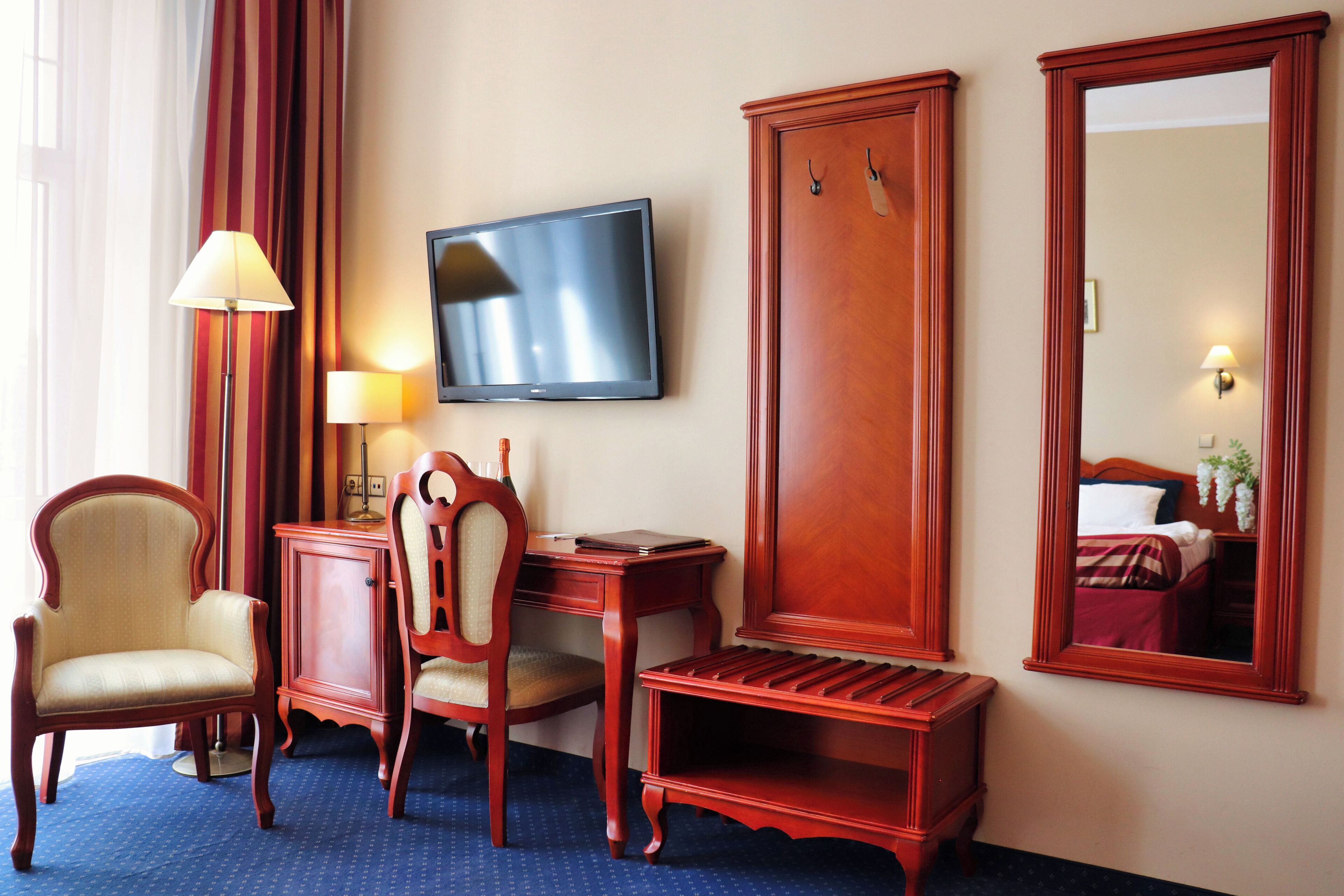 Quarto de hotel com televisão, escrivaninha com luminária, duas cadeiras, armário e um espelho grande.
