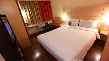 Quarto de hotel com uma cama grande, uma escrivaninha com cadeira, televisão e janela com cortinas.