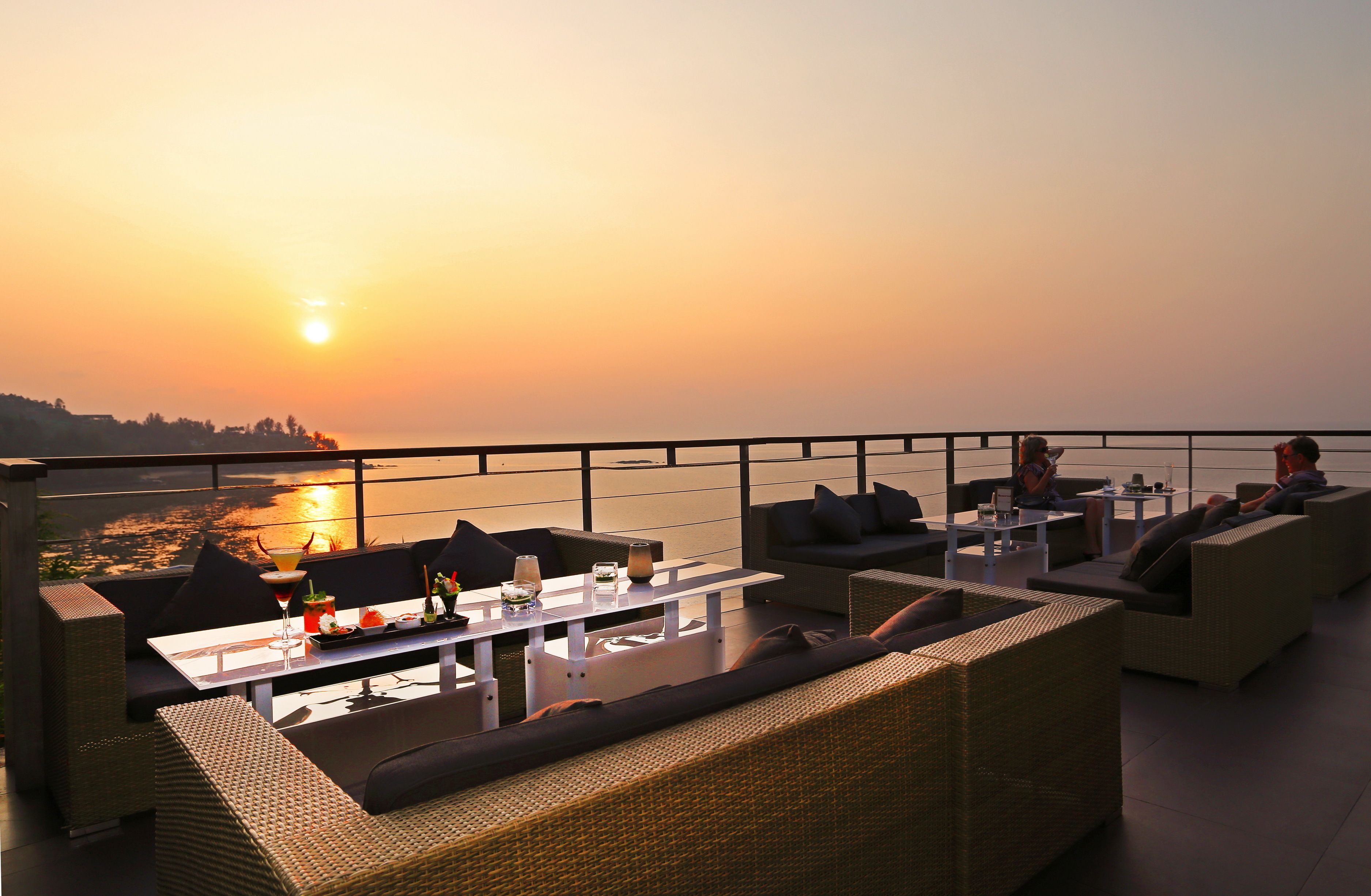 Terraço no terraço com móveis de vime e uma mesa posta para refeições, com vista para um pôr do sol sobre a água.